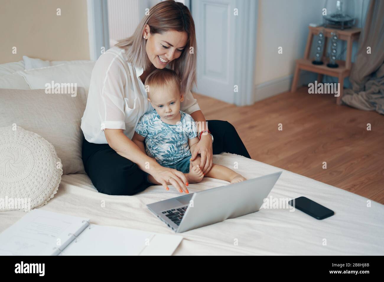 Young mother with her child working on laptop in bedroom at home. Multi-tasking, freelance and ...