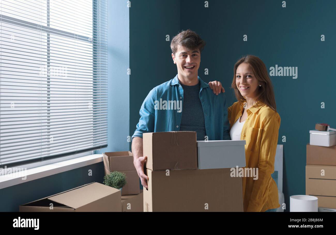 Happy young couple posing in their new house and holding carboard boxes: home relocation concept ...