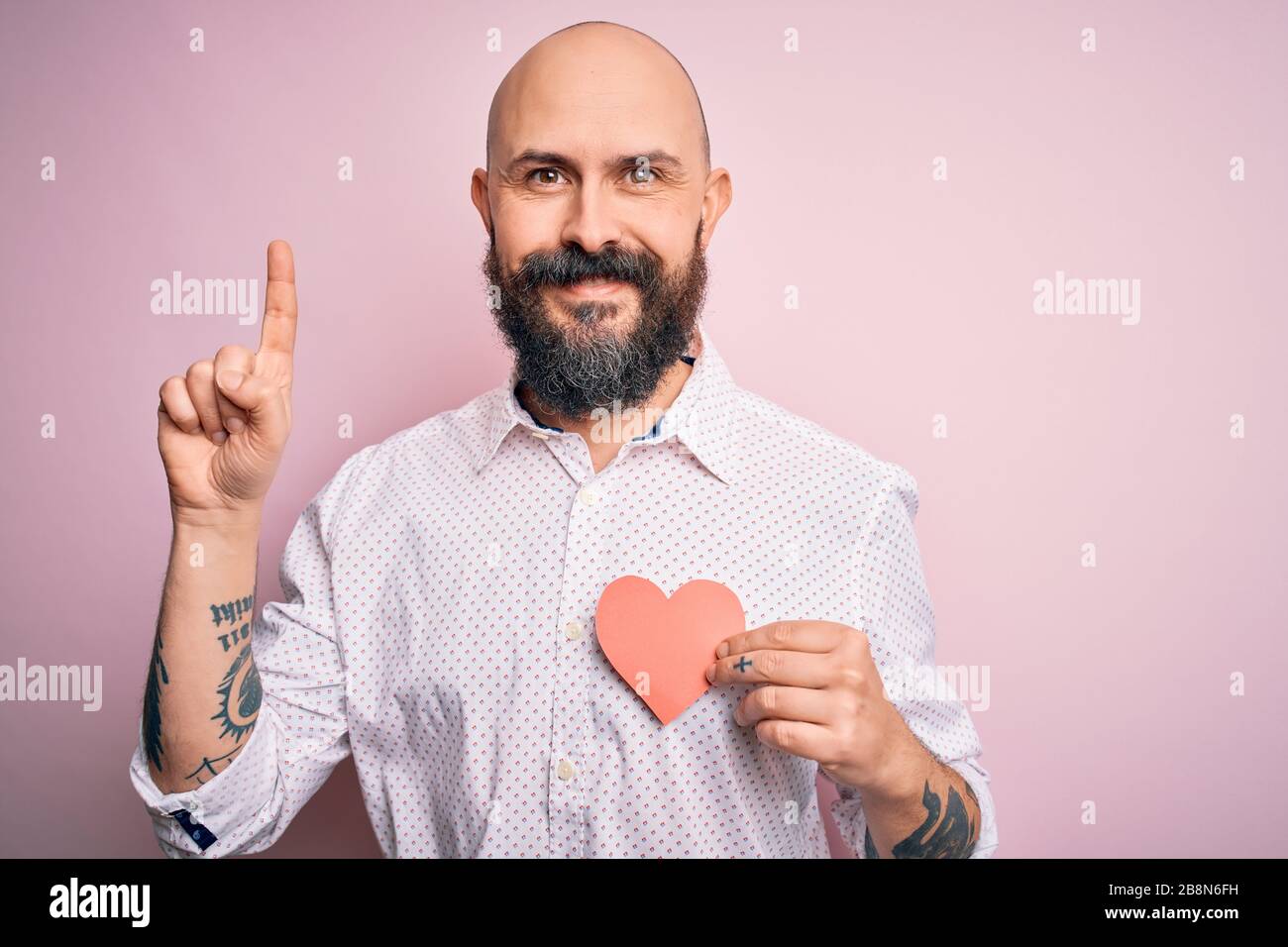 Handsome romantic bald man with beard holding red heart paper over pink ...