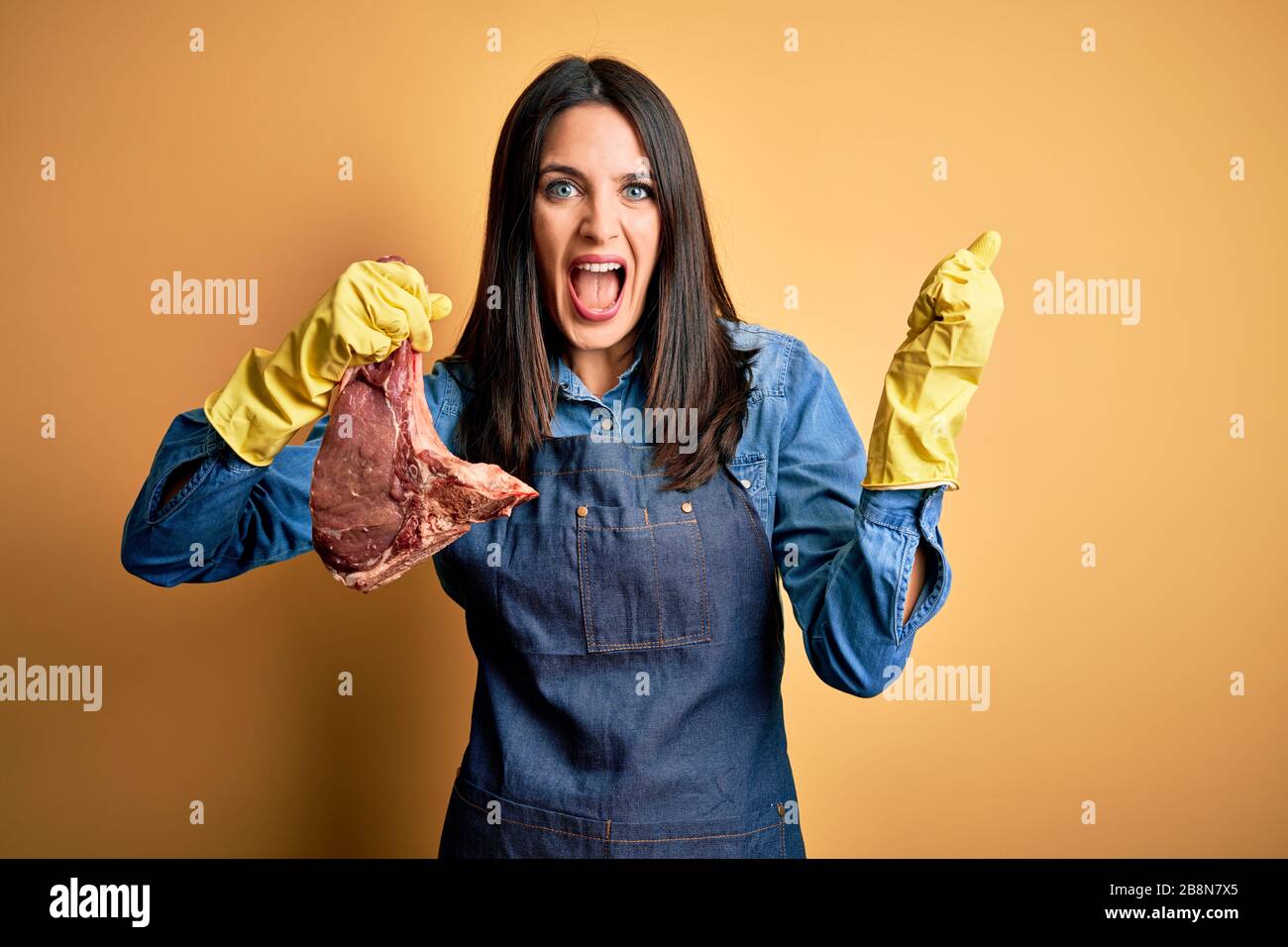 Young butcher woman wearing apron holding raw beef steak over yellow ...