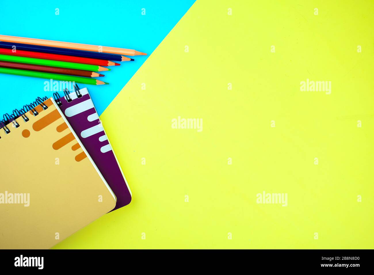 Six wood pencil crayons and two notebook scattered on a yellow and blue ...