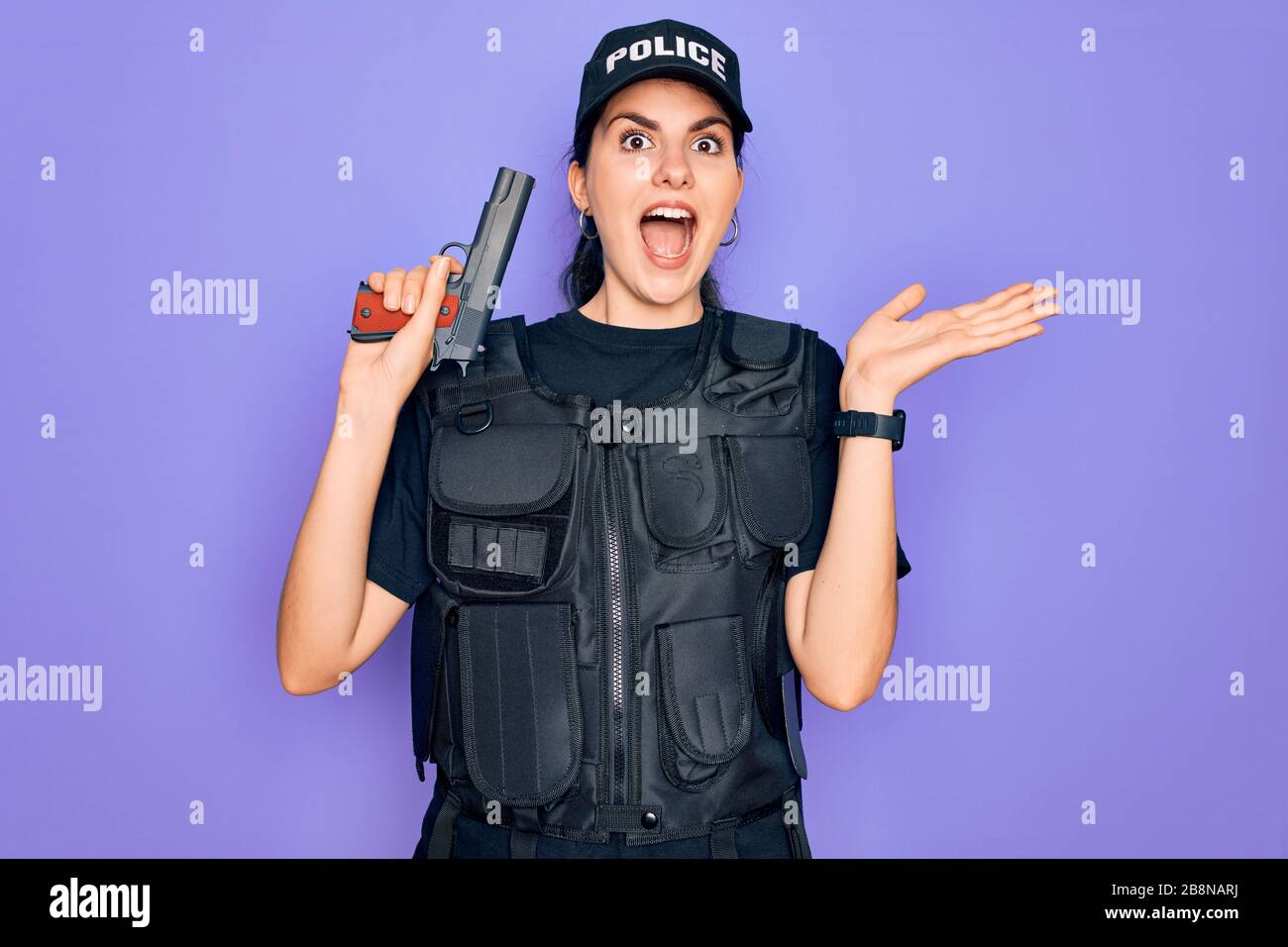 Young police woman wearing security bulletproof vest uniform and holding gun very happy and ...