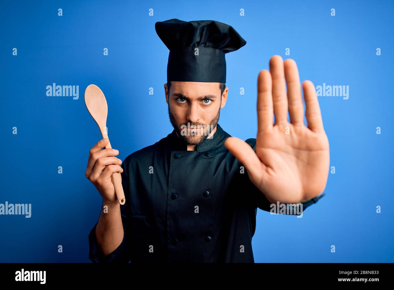 Young cooker man with beard wearing uniform holding wooden spoon over ...