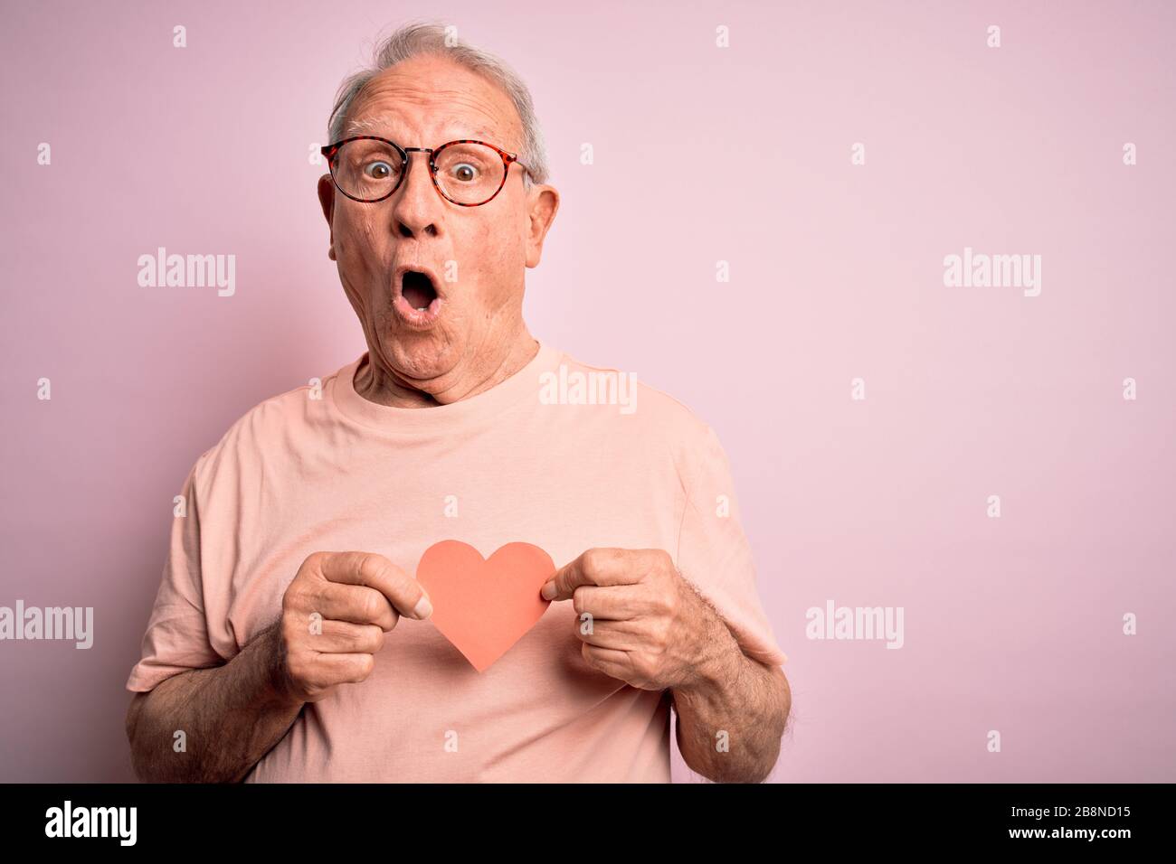 Senior grey haired man holding heart shape paper over pink background ...