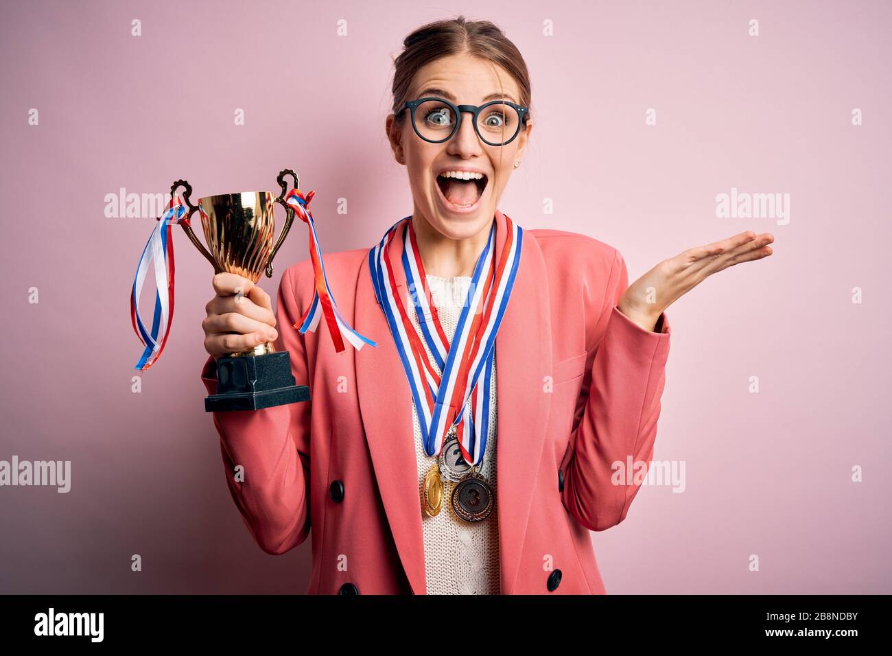 Young beautiful redhead woman holding trophy wearing medals over pink ...