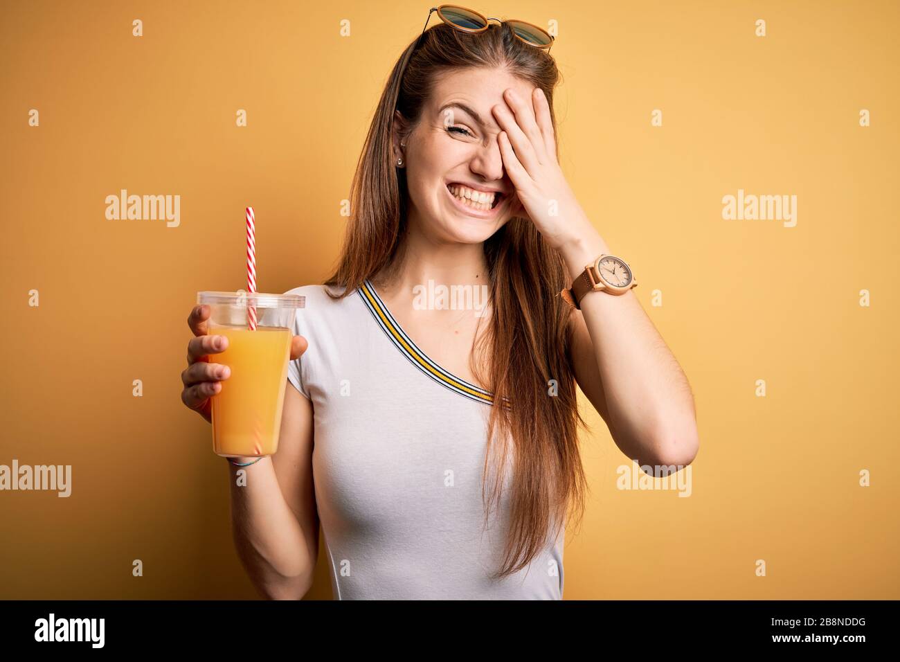 Young beautiful redhead woman drinking healthy orange juice over yellow ...