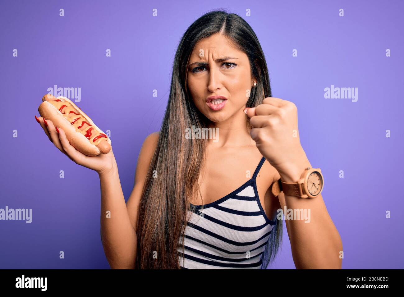 Young woman eating hotdog with ketchup and mustard over purple ...