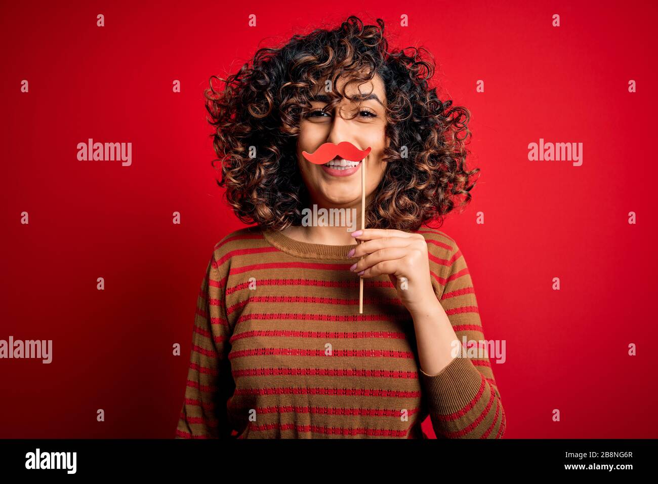 Young beautiful curly arab woman holding funny mustache over isolated ...