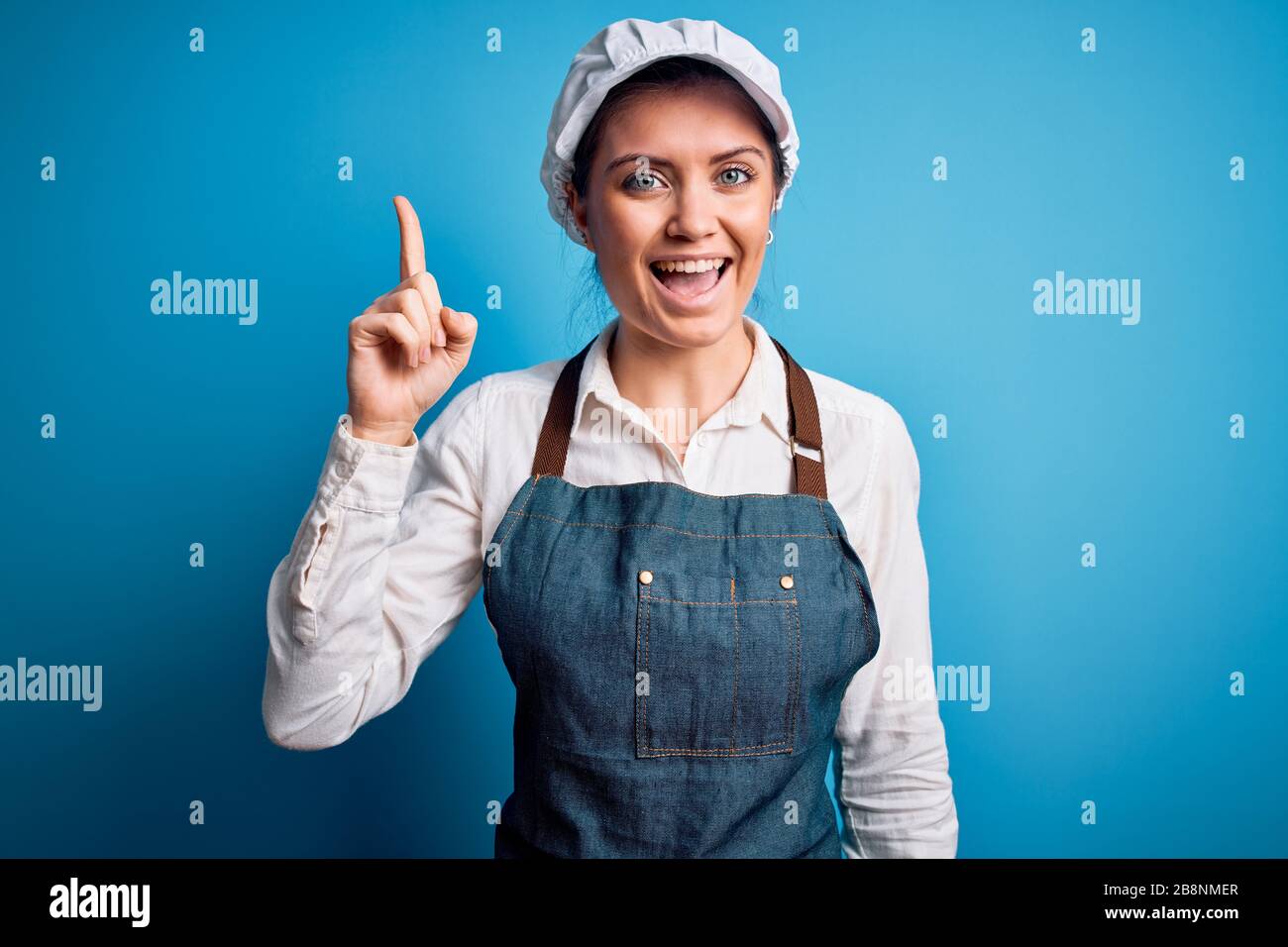 Young beautiful baker woman with blue eyes wearing apron and cap over ...
