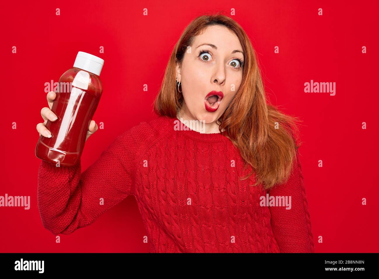 Beautiful Redhead Woman Holding Bottle Of Ketchup Sauce Condiment Over Red Background Scared In