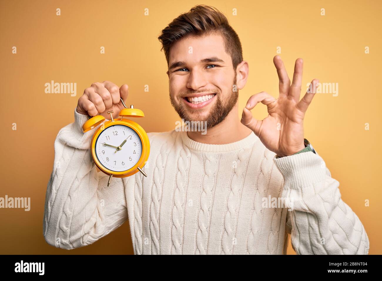 Young blond man with beard and blue eyes holding alarm clock over ...