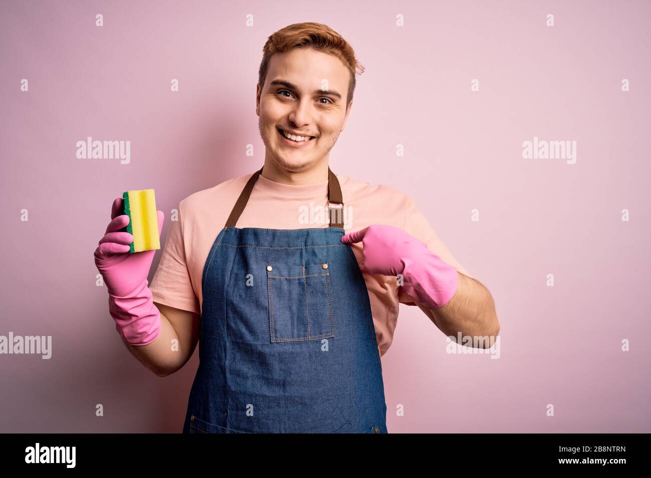 Handsome Redhead Man Doing Housework Wearing Apron And Gloves Using Cleaner Scourer Pointing
