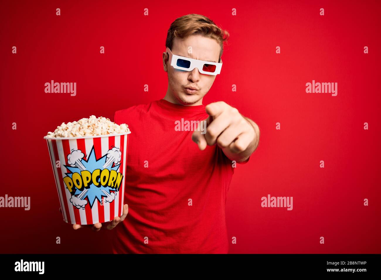 Young handsome redhead man watching 3d glasses eating popcorn snack over red background pointing ...