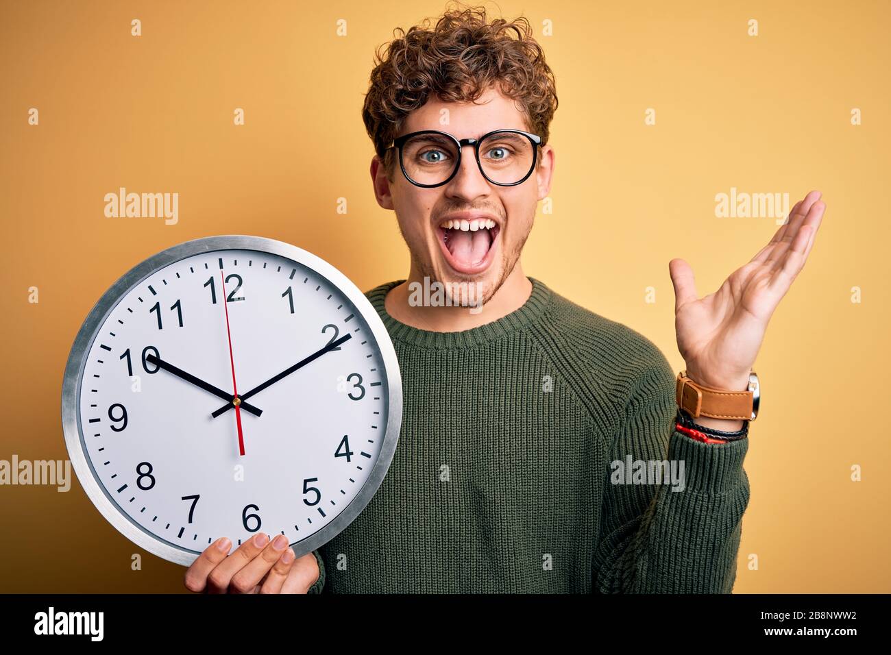 Young blond man with curly hair wearing glasses holding big clock over ...