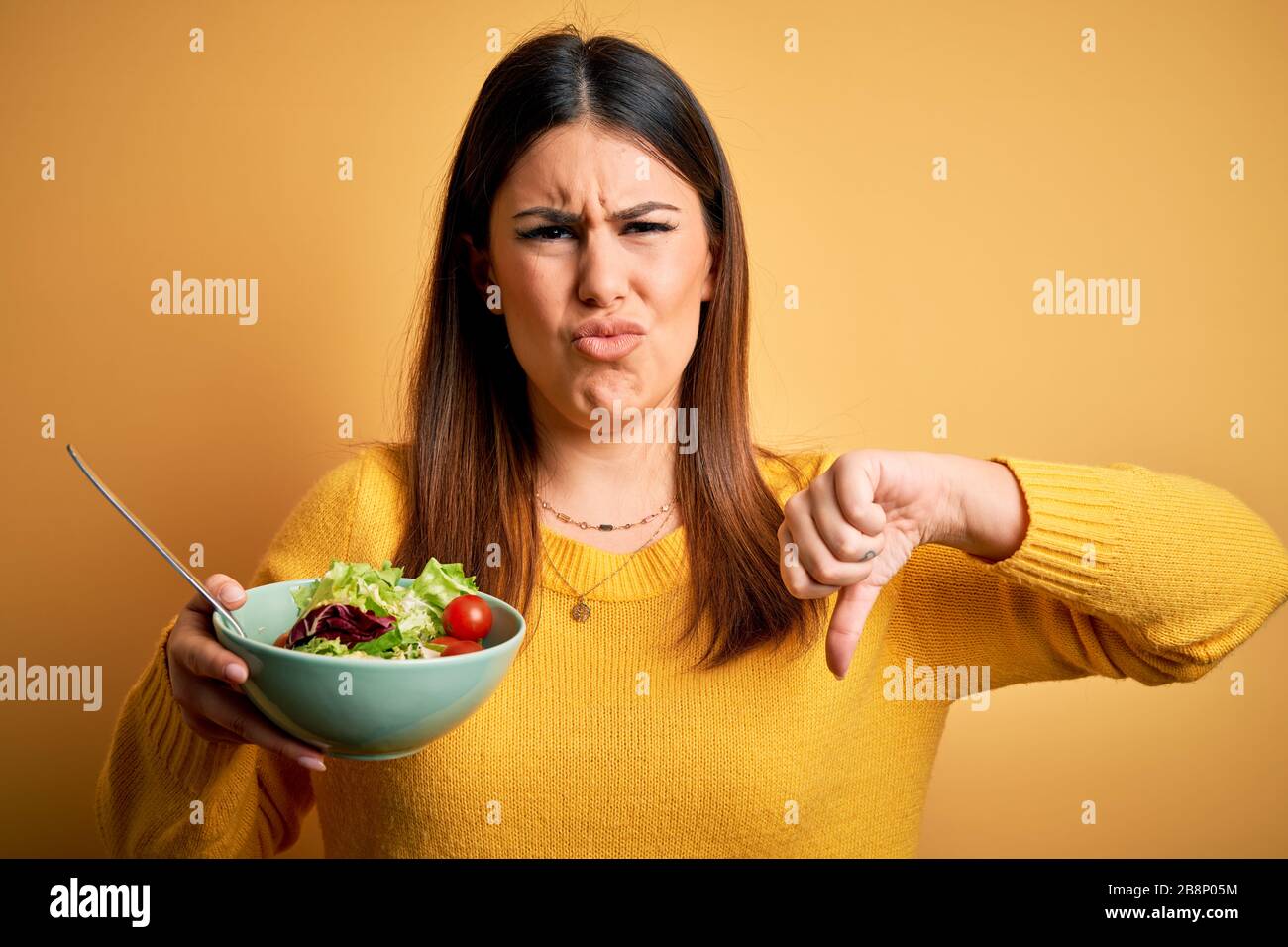 Young beautiful woman eating healthy fresh salad over yellow background ...