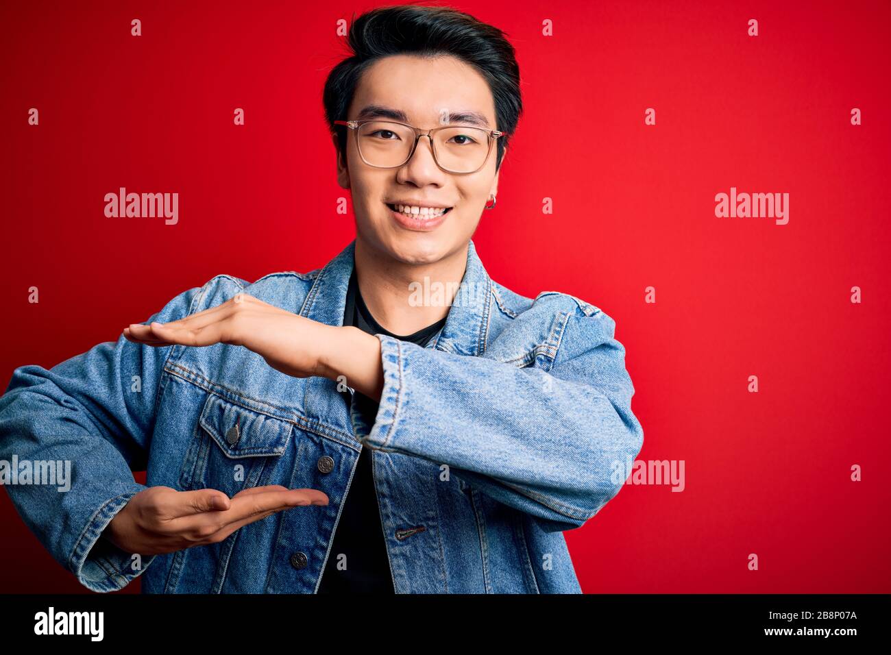 Young handsome chinese man wearing denim jacket and glasses over red ...