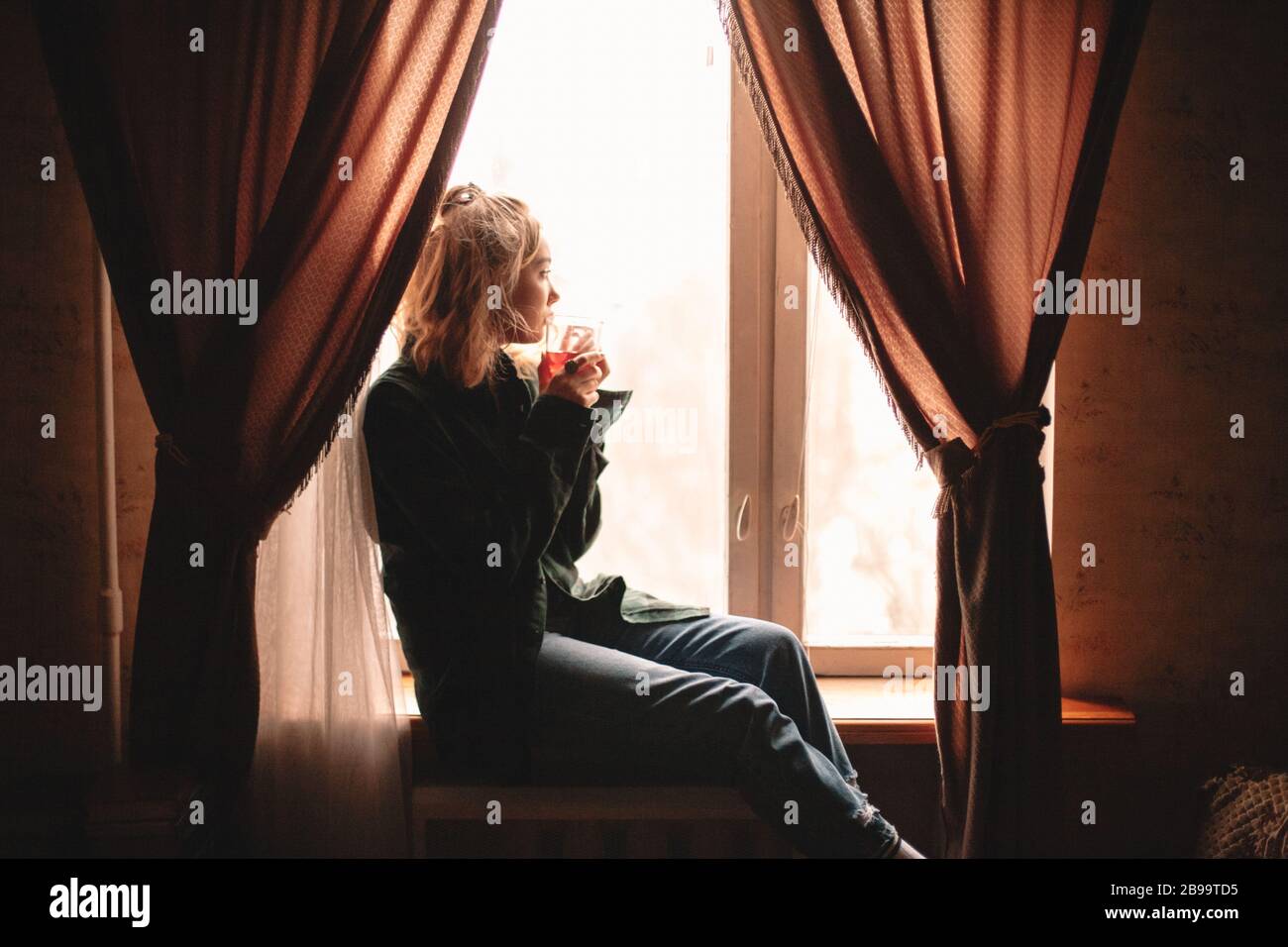 Young thoughtful woman drinking tea and looking through window while ...