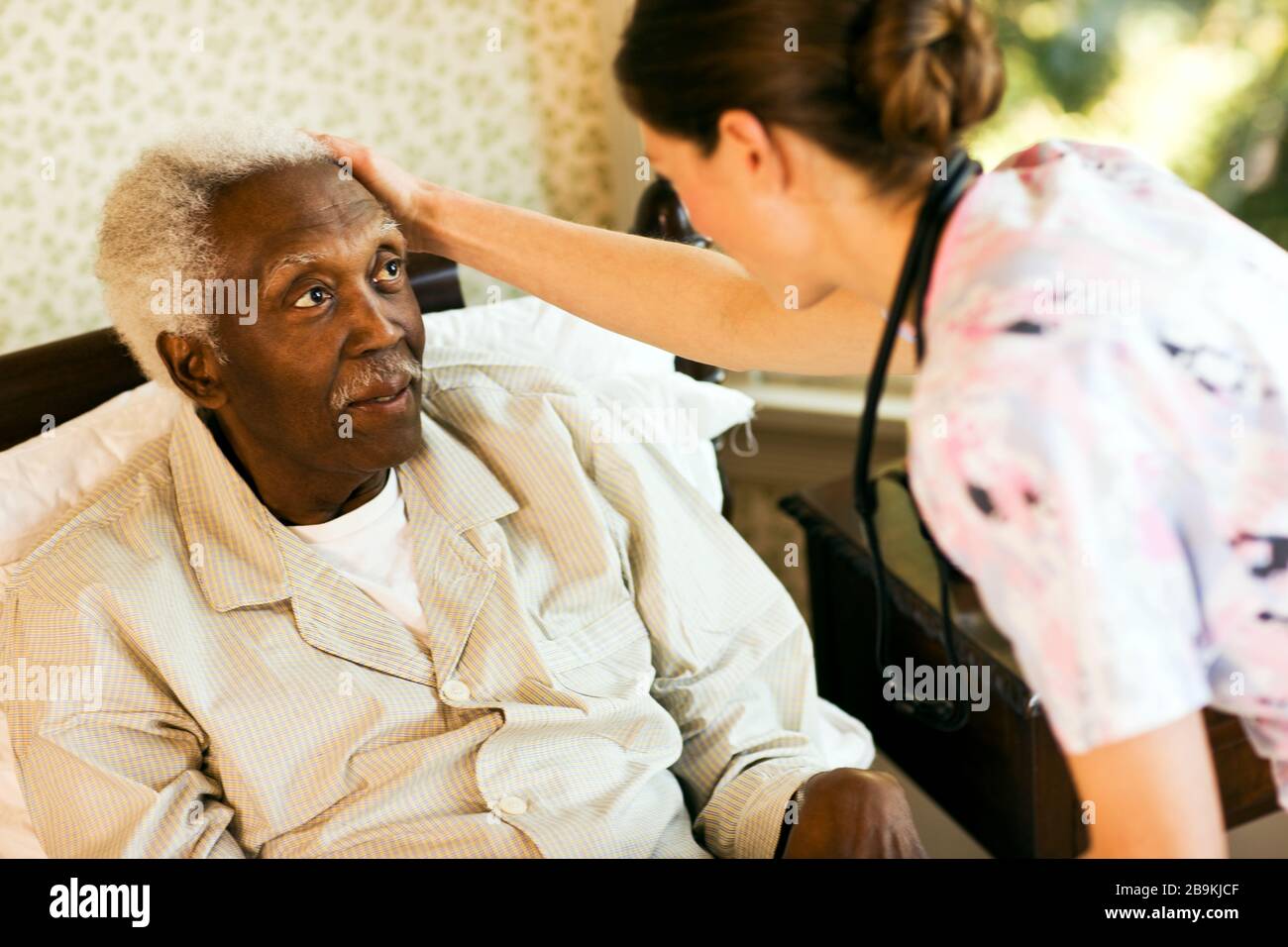 Senior man sitting in bed looks up at a female nurse who leans over to ...