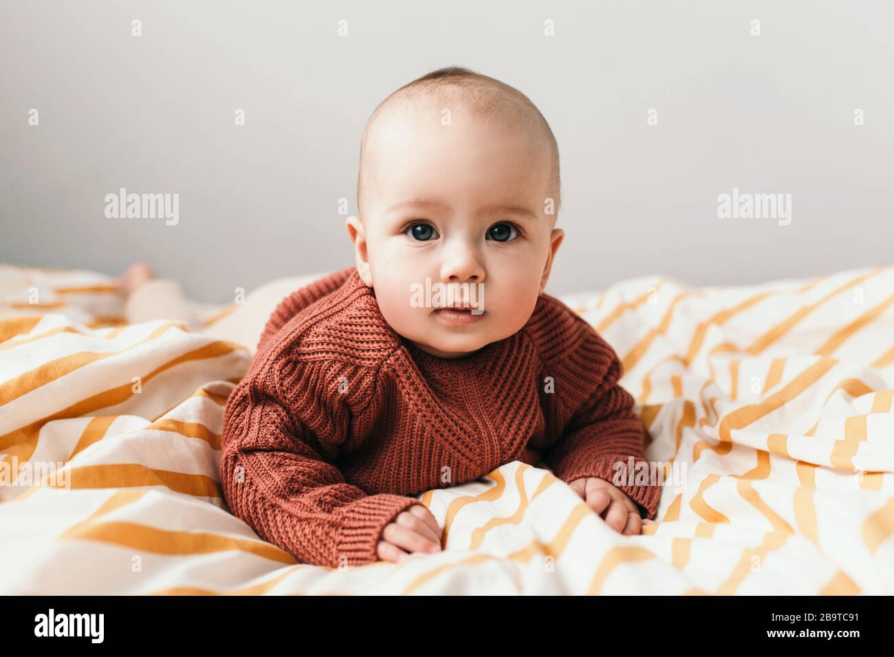 Beautiful little baby girl on the bed in a cozy brown sweater smiles. Concept of motherhood and ...