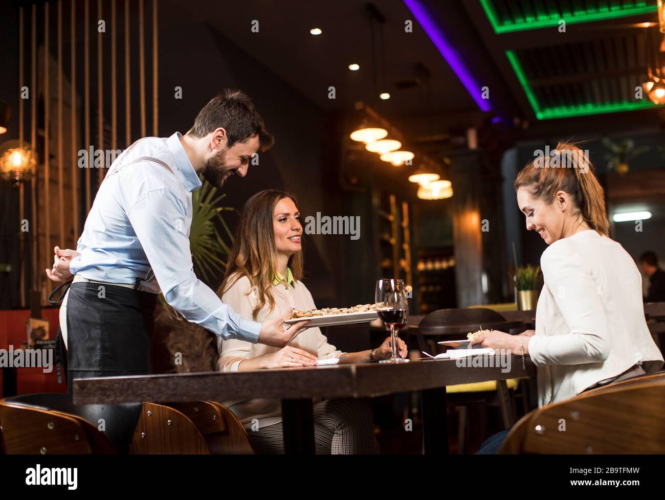 Two smiling young female friends at a restaurant with waiter serving ...