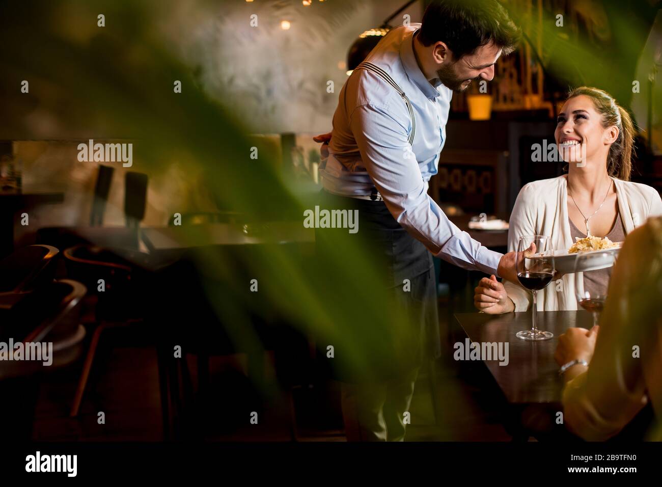 Two smiling young female friends at a restaurant with waiter serving ...