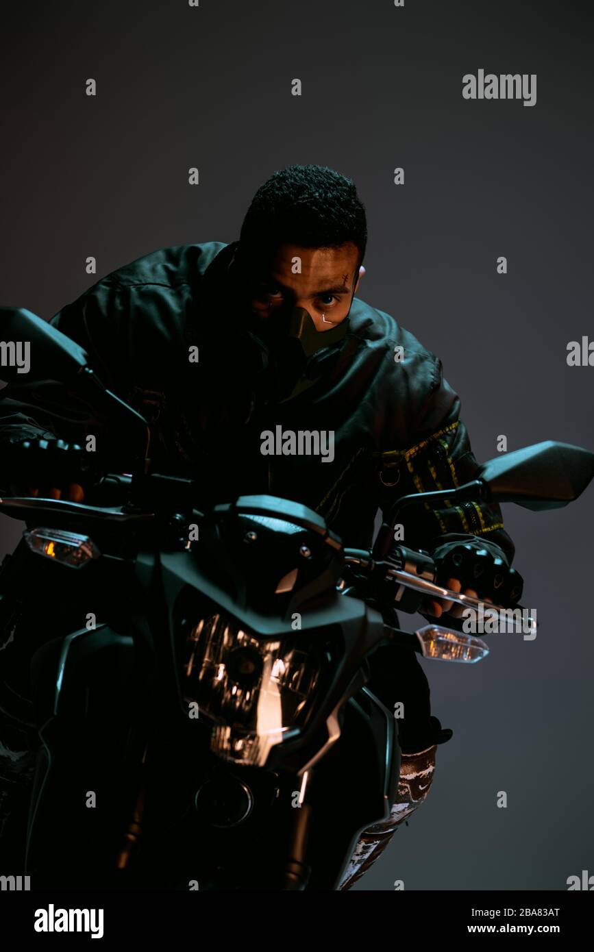 handsome mixed race cyberpunk player in mask riding motorcycle on grey ...