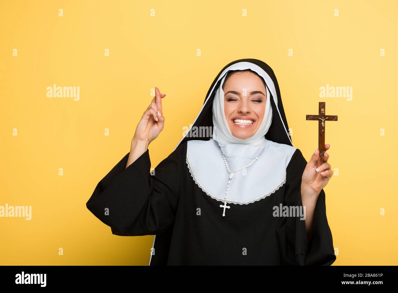 cheerful nun holding cross while showing fingers crossed on yellow ...
