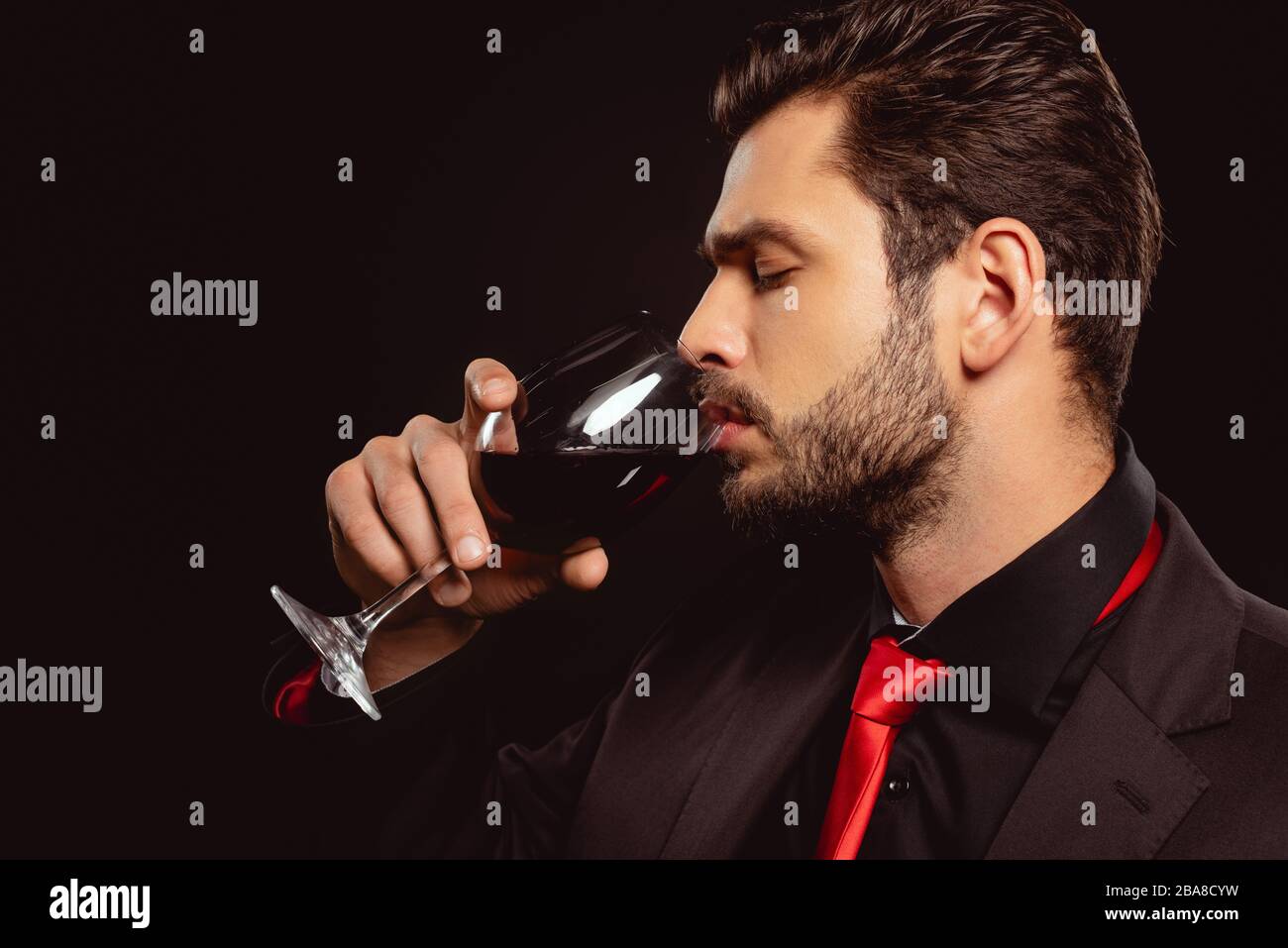 Side view of handsome man in suit drinking red wine isolated on black ...