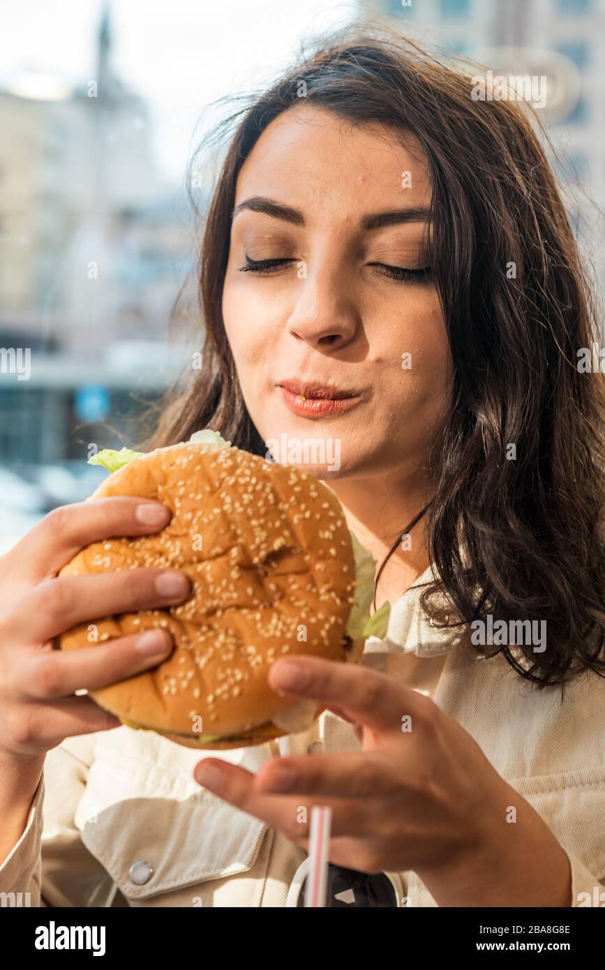 Beautiful young cute girl in fashionable clothes enjoys a big fast food ...