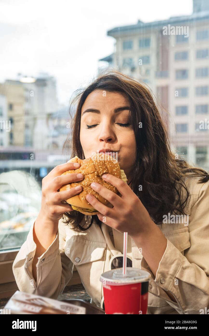 Beautiful young cute girl in fashionable clothes enjoys a big fast food ...