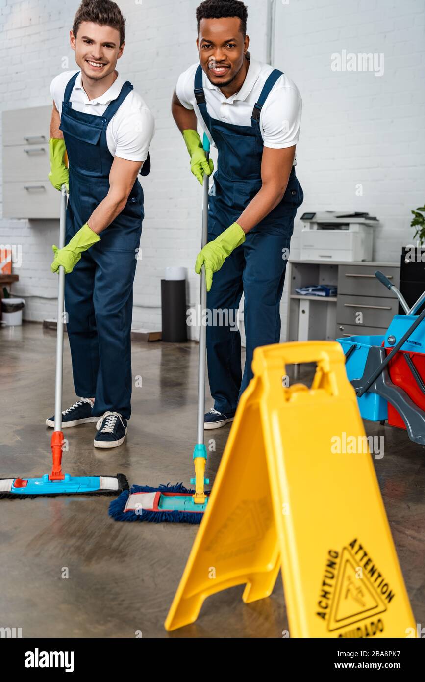 smiling multicultural cleaners washing floor with mops near wet floor ...