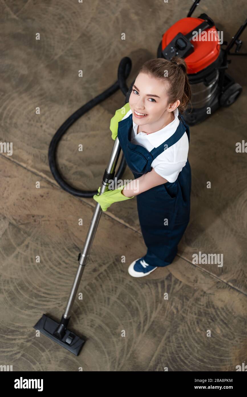 overhead view of smiling cleaner looking at camera while vacuuming ...