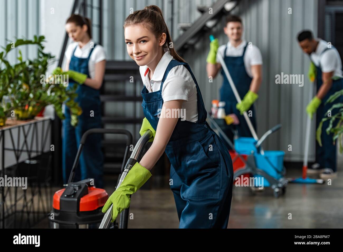 smiling cleaner vacuuming floor in office near multicultural colleagues ...