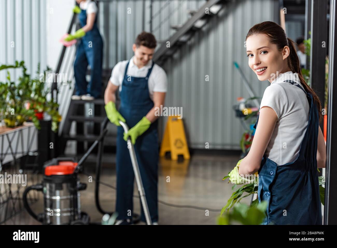 attractive cleaner smiling at camera while colleagues cleaning office ...