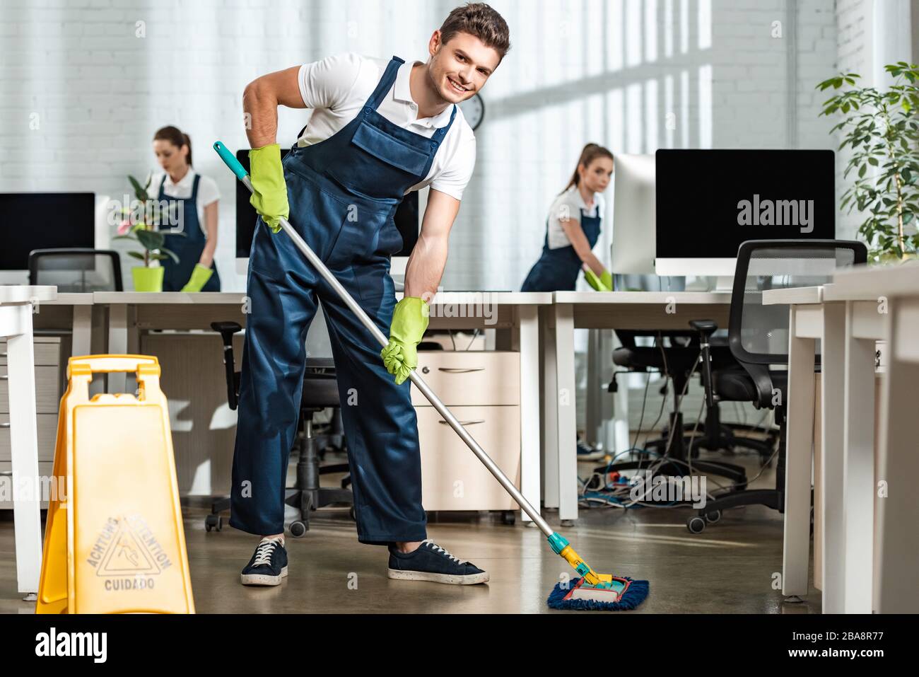 young cleaner looking at camera while washing floor with mop in modern ...