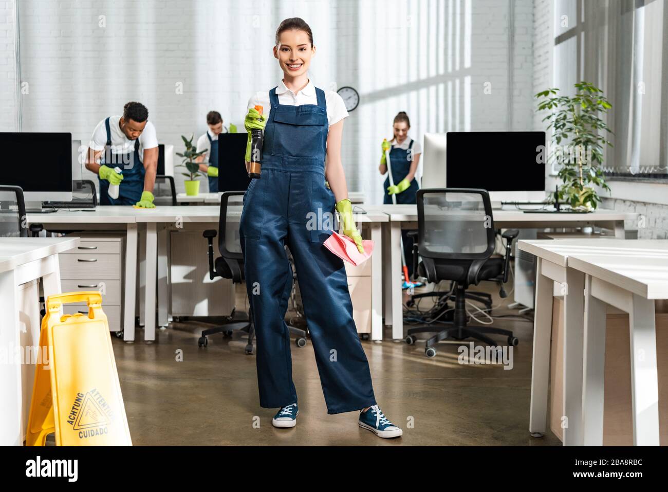 smiling cleaner holding spray and rag while standing near working ...