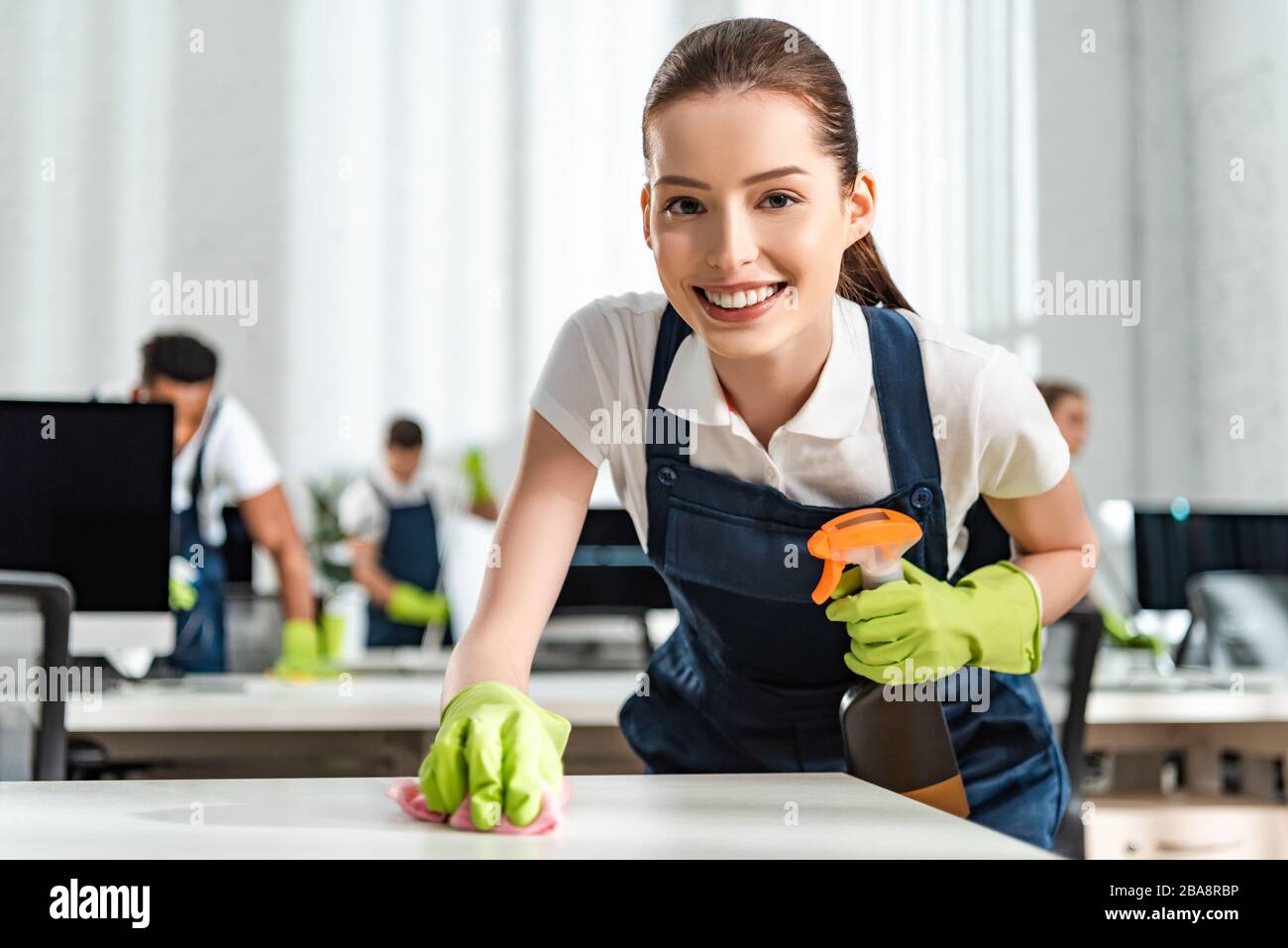 happy cleaner looking at camera while cleaning office desk with rag ...