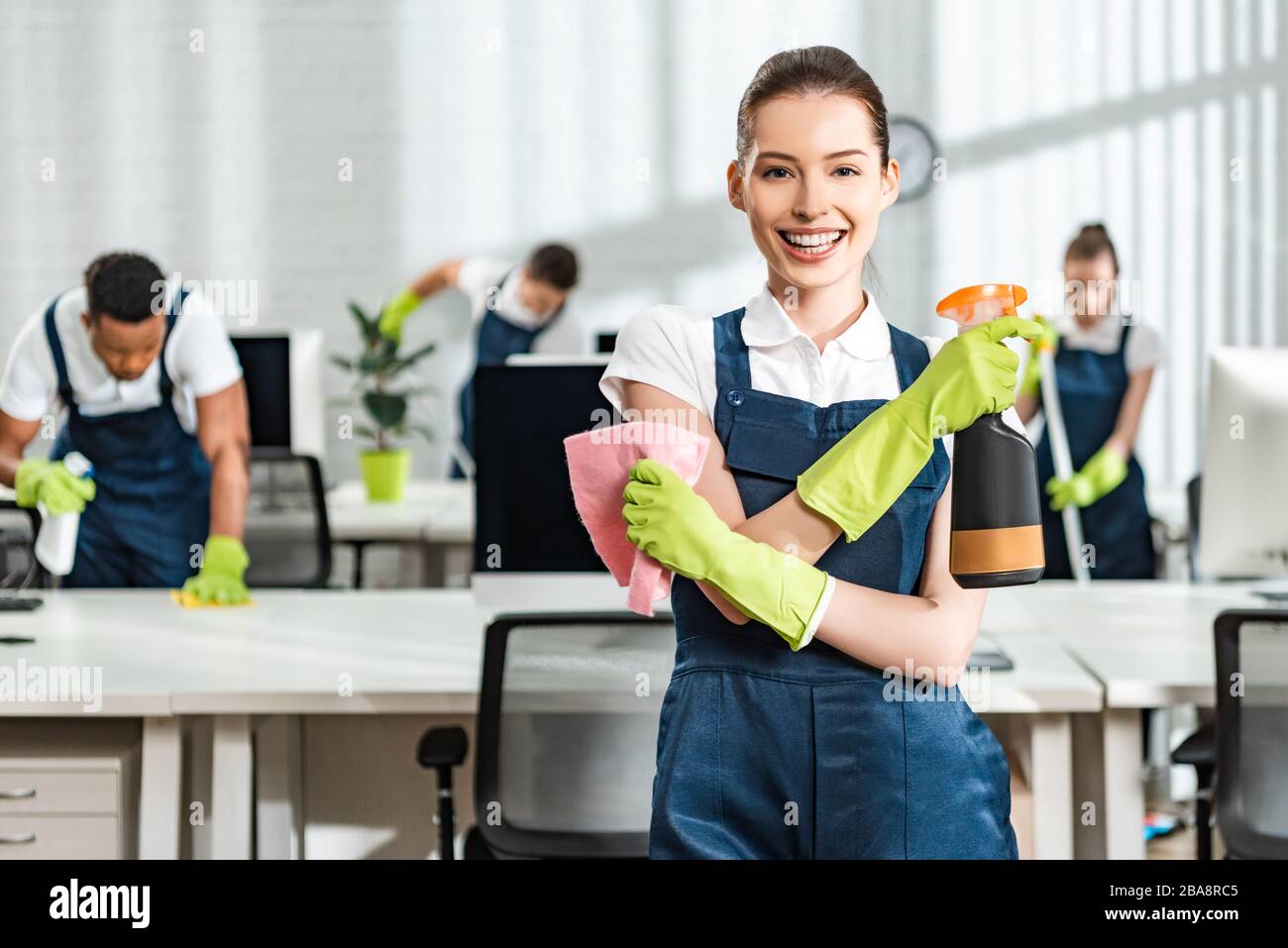 happy cleaner with spray and rag smiling at camera while standing near ...