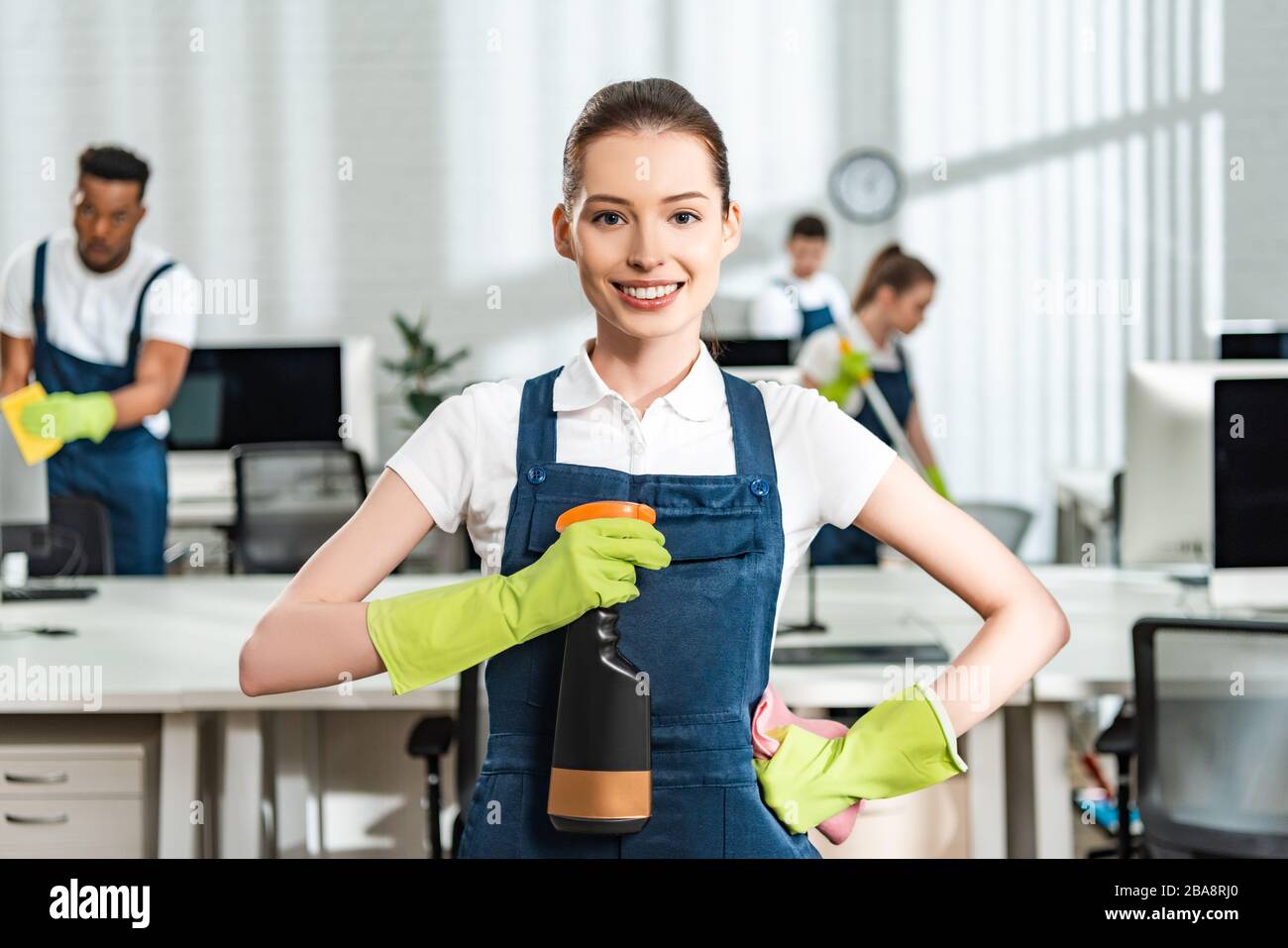 happy cleaner standing with hand on hip and holding spray bottle while ...