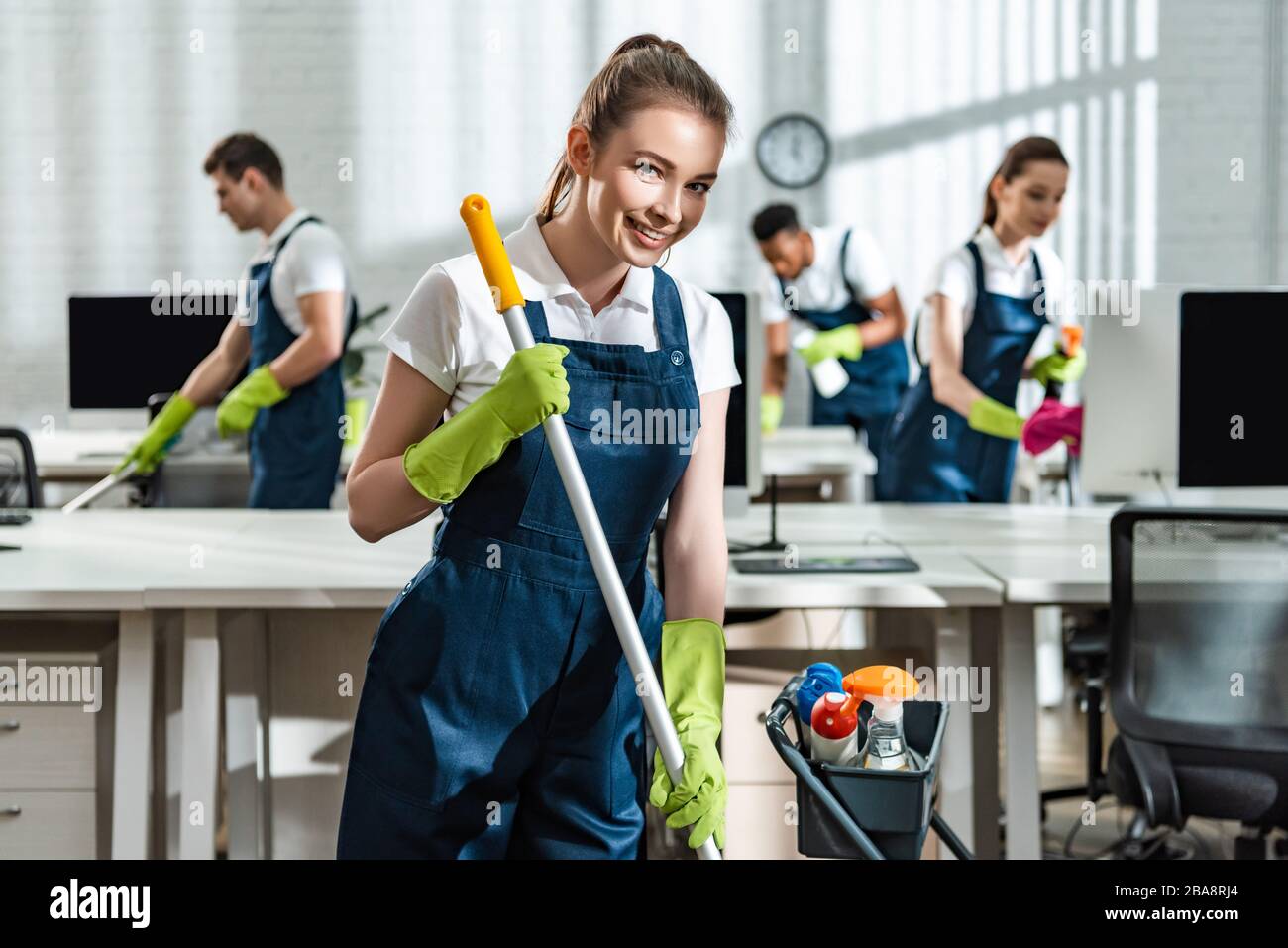 smiling cleaner washing floor and smiling at camera near multicultural ...