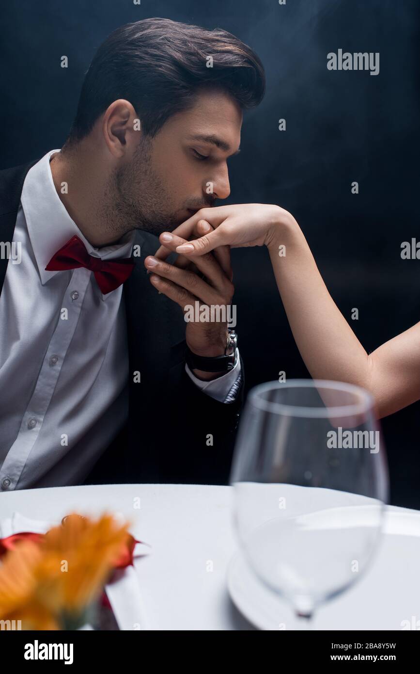 Side view of man in suit kissing hand of woman at served table on black ...