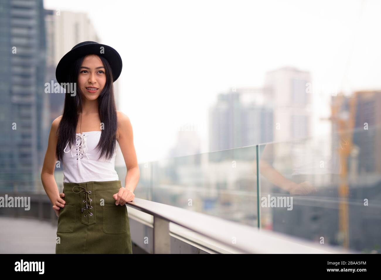 Happy young beautiful Asian tourist woman thinking against view of the ...