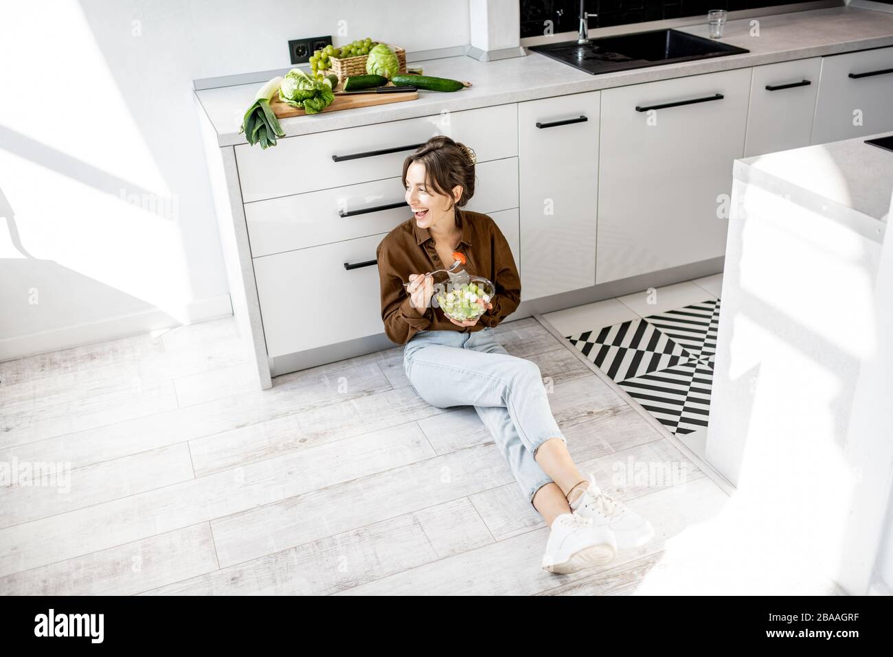 Young woman sitting on the white kitchen floor, eating salad while ...