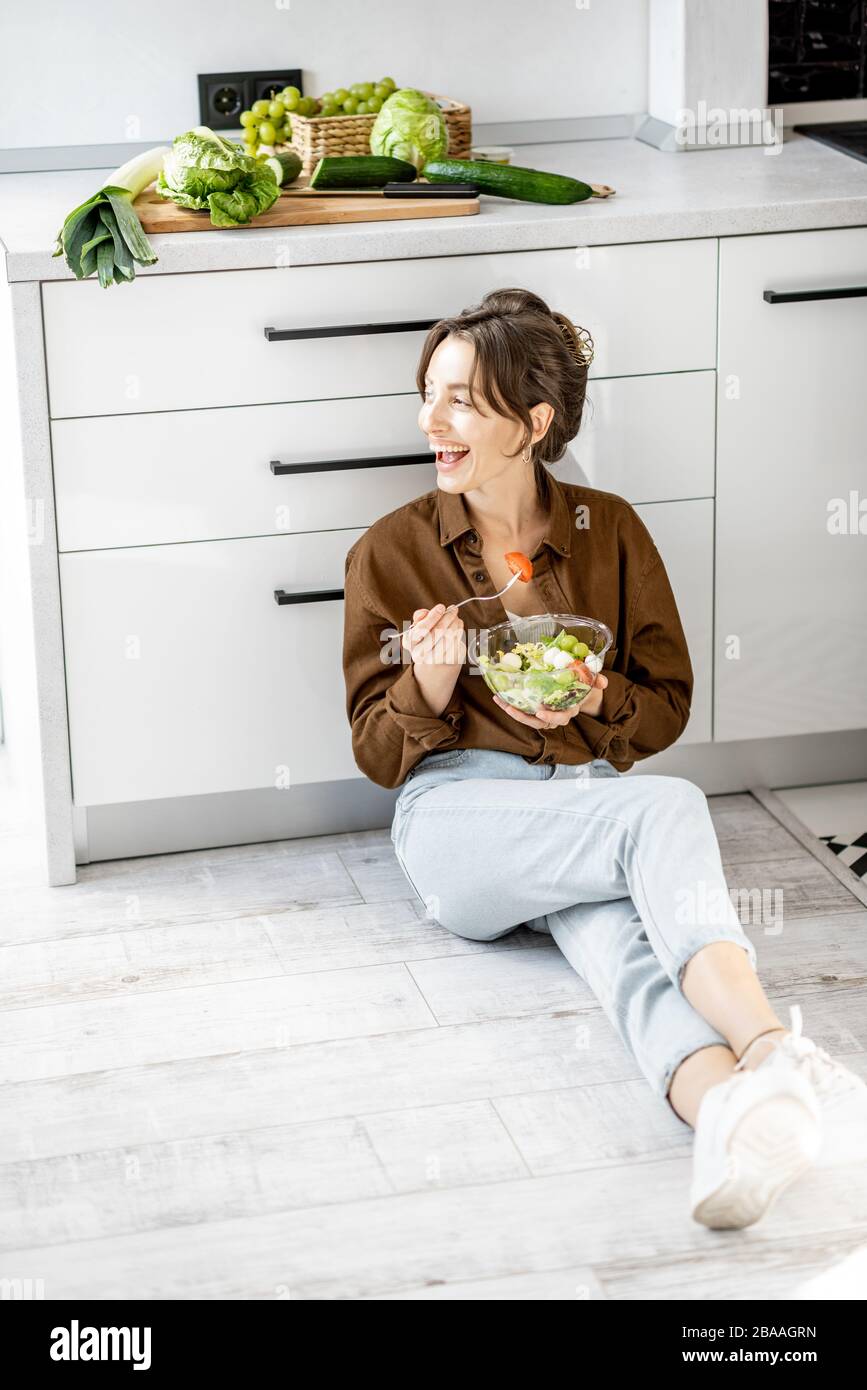 Young woman sitting on the white kitchen floor, eating salad while ...