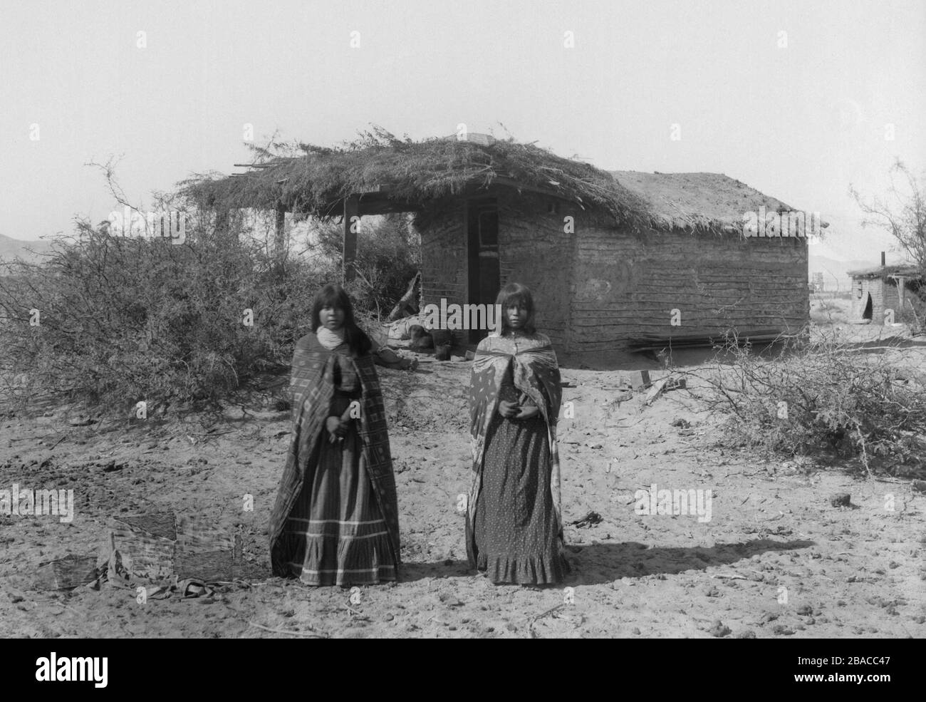 Two Mojave Indians girls stand in front of a small wattle and daub ...