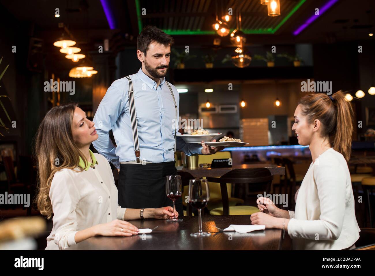 Two smiling young female friends at a restaurant with waiter serving ...