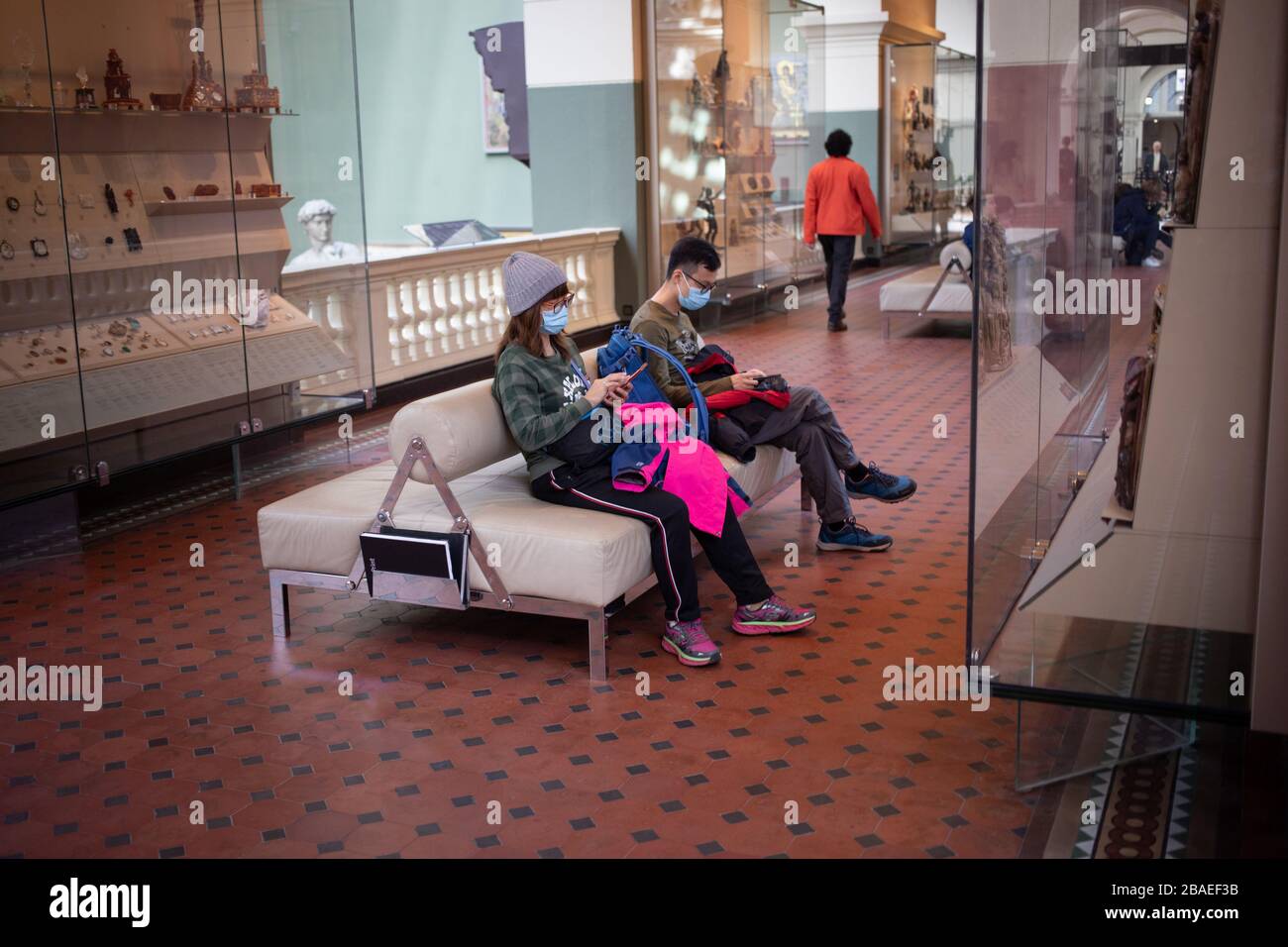 Two visitors to the V&A museum sit on a sofa and look at their mobile ...
