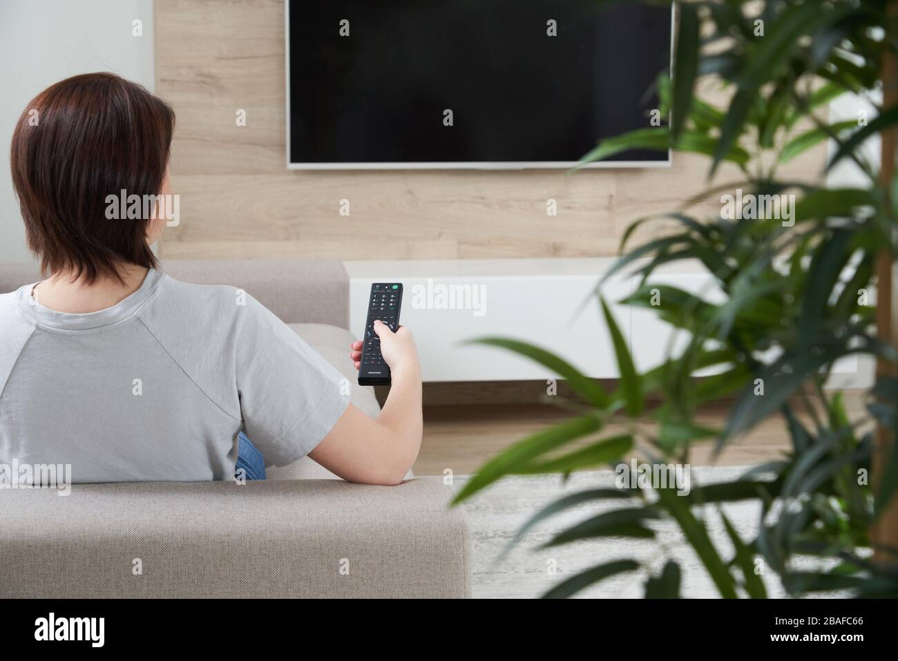 Rear view of young woman watching television and holding remote control ...