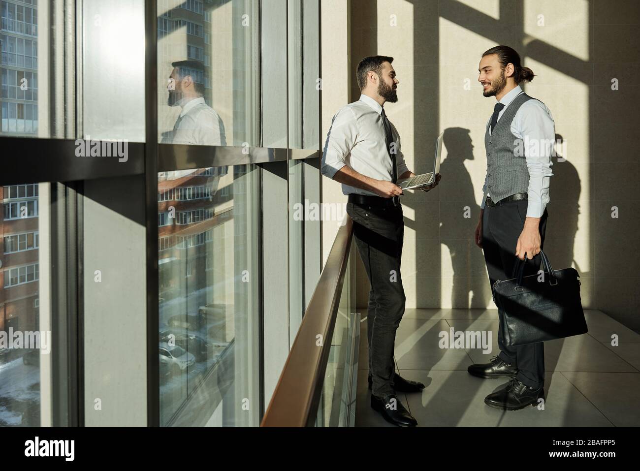 Two young successful businessmen standing by large window during ...