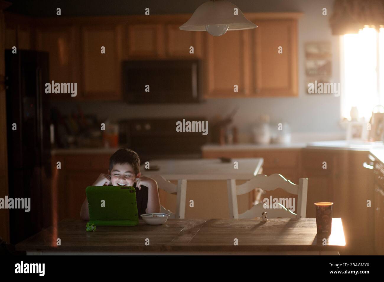 Young boy laughing while watching tablet at kitchen table eating food ...